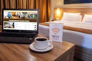 Um laptop exibindo uma página de reserva de hotel, uma xícara de café e um sinal indicando acesso à rede Wi-Fi.