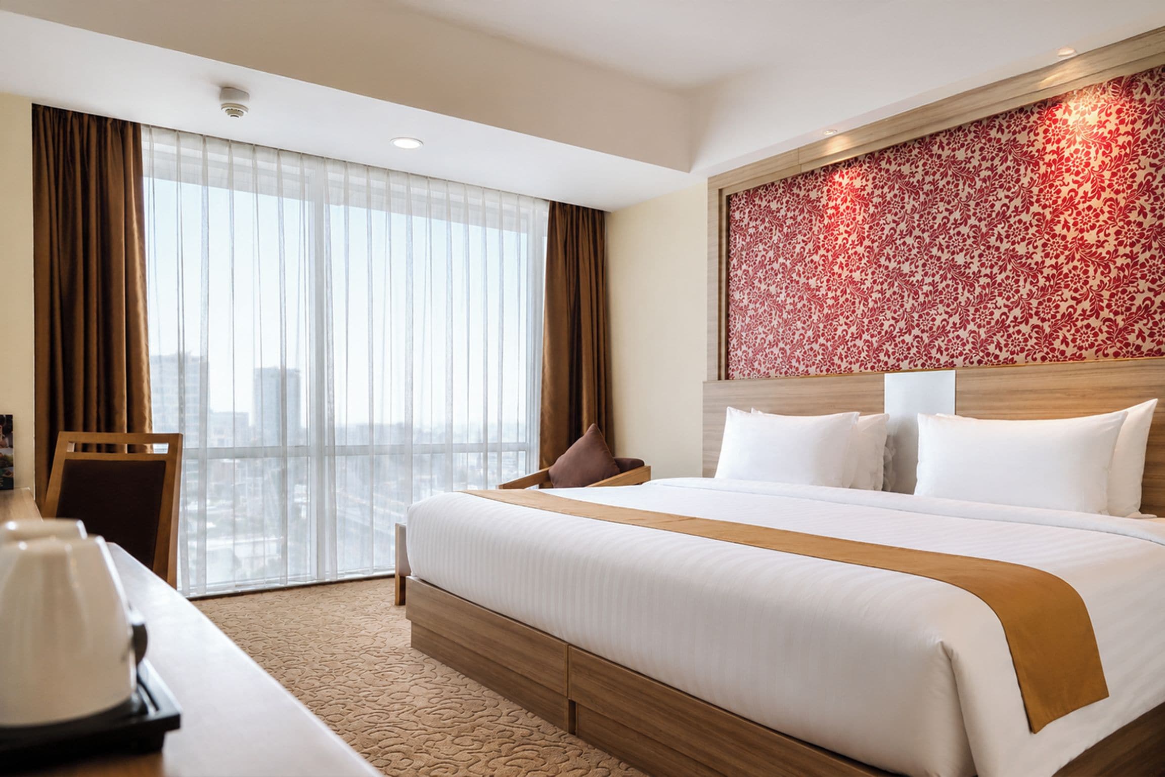 Quarto de hotel com uma cama grande, uma mesa com cadeira, vista para a cidade pela janela e um painel decorativo na parede.