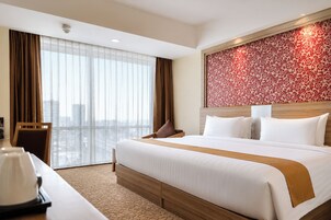 Quarto de hotel com uma cama grande, uma mesa com cadeira, vista para a cidade pela janela e um painel decorativo na parede.