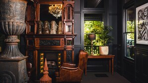 Quarto com uma lareira de madeira grande e ornamentada, uma poltrona de couro e um vaso de plantas perto de uma janela.