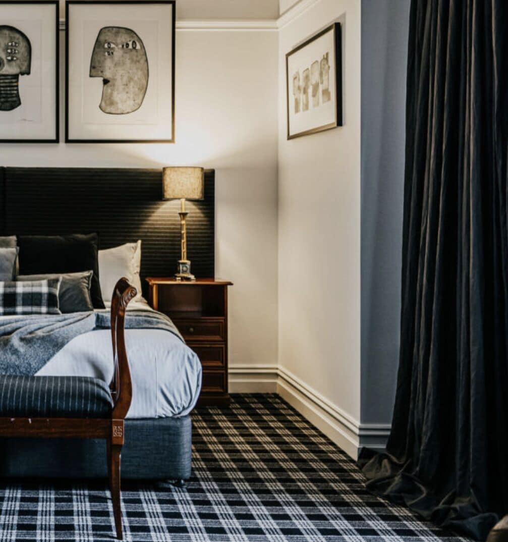 Quarto com piso xadrez, cama com cabeceira de madeira, mesa de cabeceira com abajur e quadros na parede.