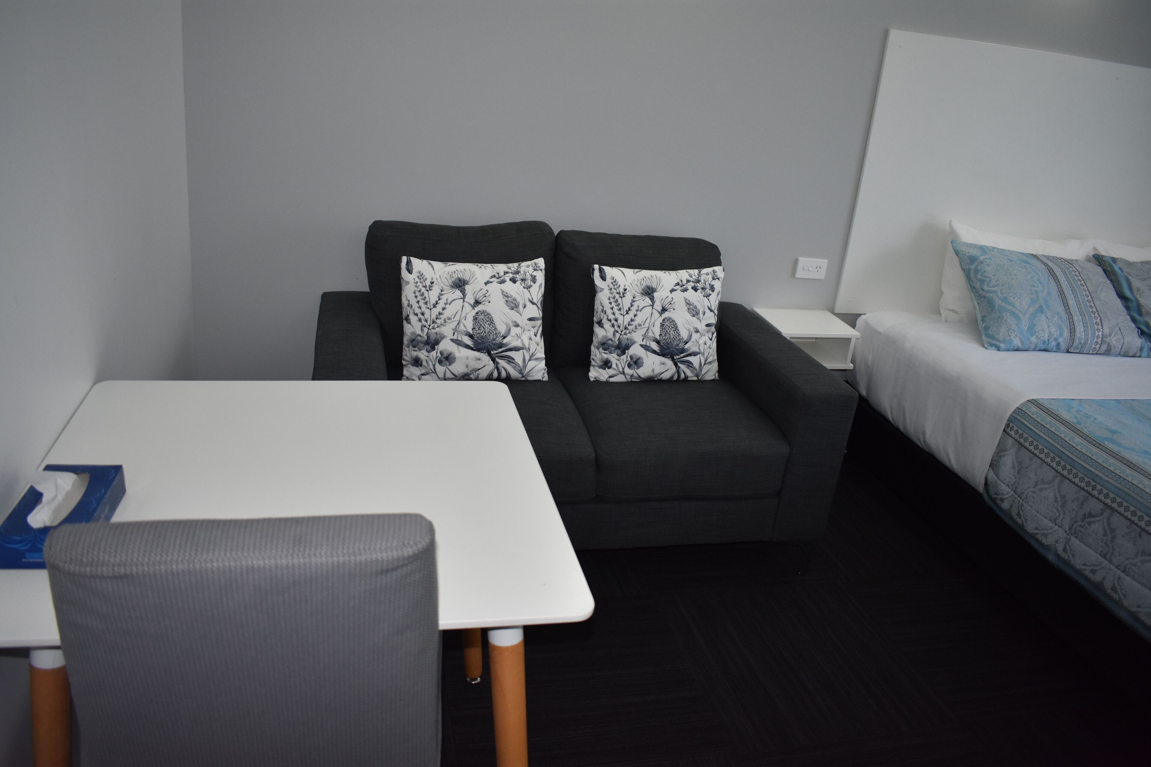 Quarto com sofá cinzento, mesa branca, cama com roupa de cama estampada e mesa de cabeceira com abajur.