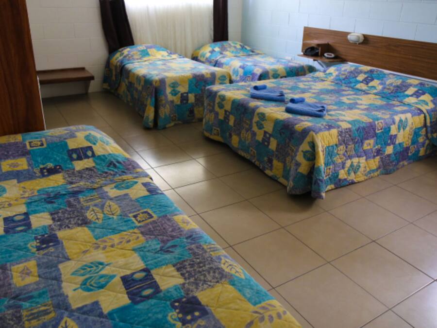Quarto com três camas, cada uma com colcha estampada. Há um banco encostado na parede e uma cabeceira de madeira em uma das camas.