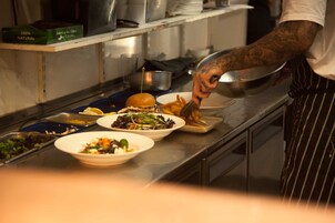 Um chef servindo comida em uma cozinha com vários pratos e ingredientes sobre o balcão.
