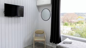 Quarto com televisão montada, espelho redondo, cadeira, cama com toalha branca dobrada e janela com vista para vegetação e edifícios.