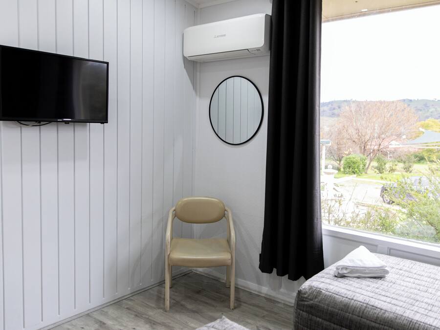 Quarto com televisão montada, espelho redondo, cadeira, cama com toalha branca dobrada e janela com vista para vegetação e edifícios.