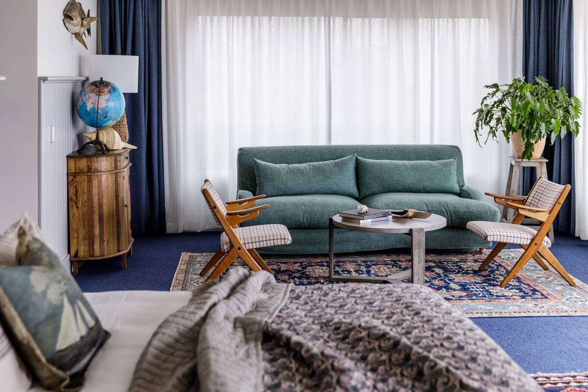 Uma sala de estar com um sofá verde, duas cadeiras de madeira, um globo em um armário de madeira, uma planta em vaso e um tapete estampado.