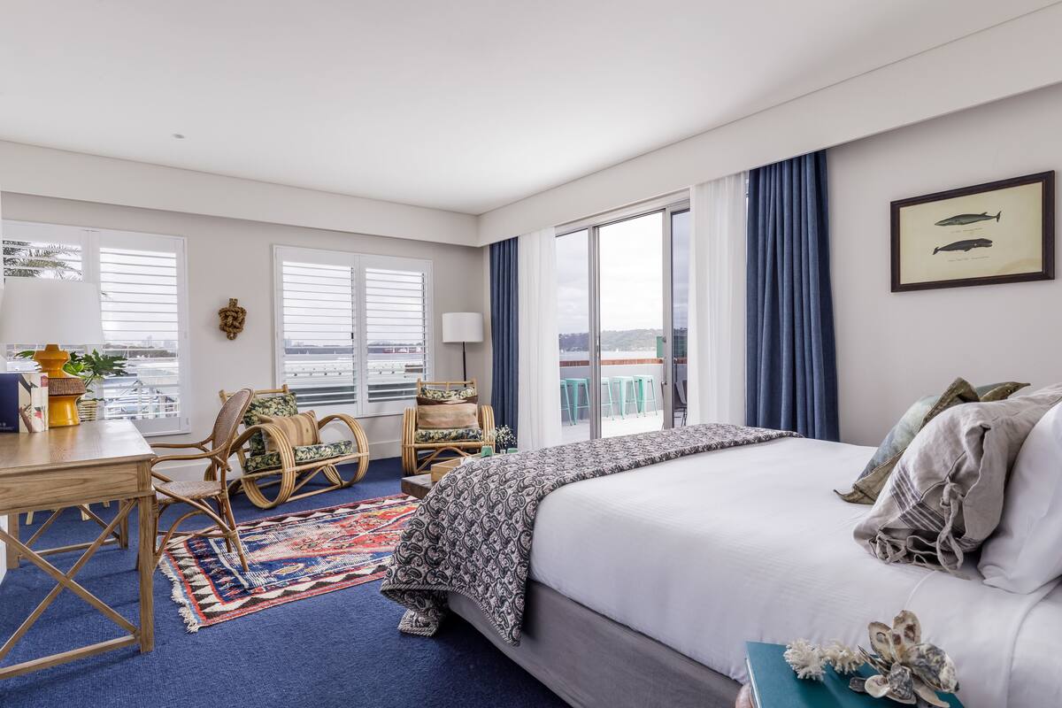 Quarto de hotel com uma cama grande, uma mesa, uma cadeira e vista da cidade através de janelas amplas.
