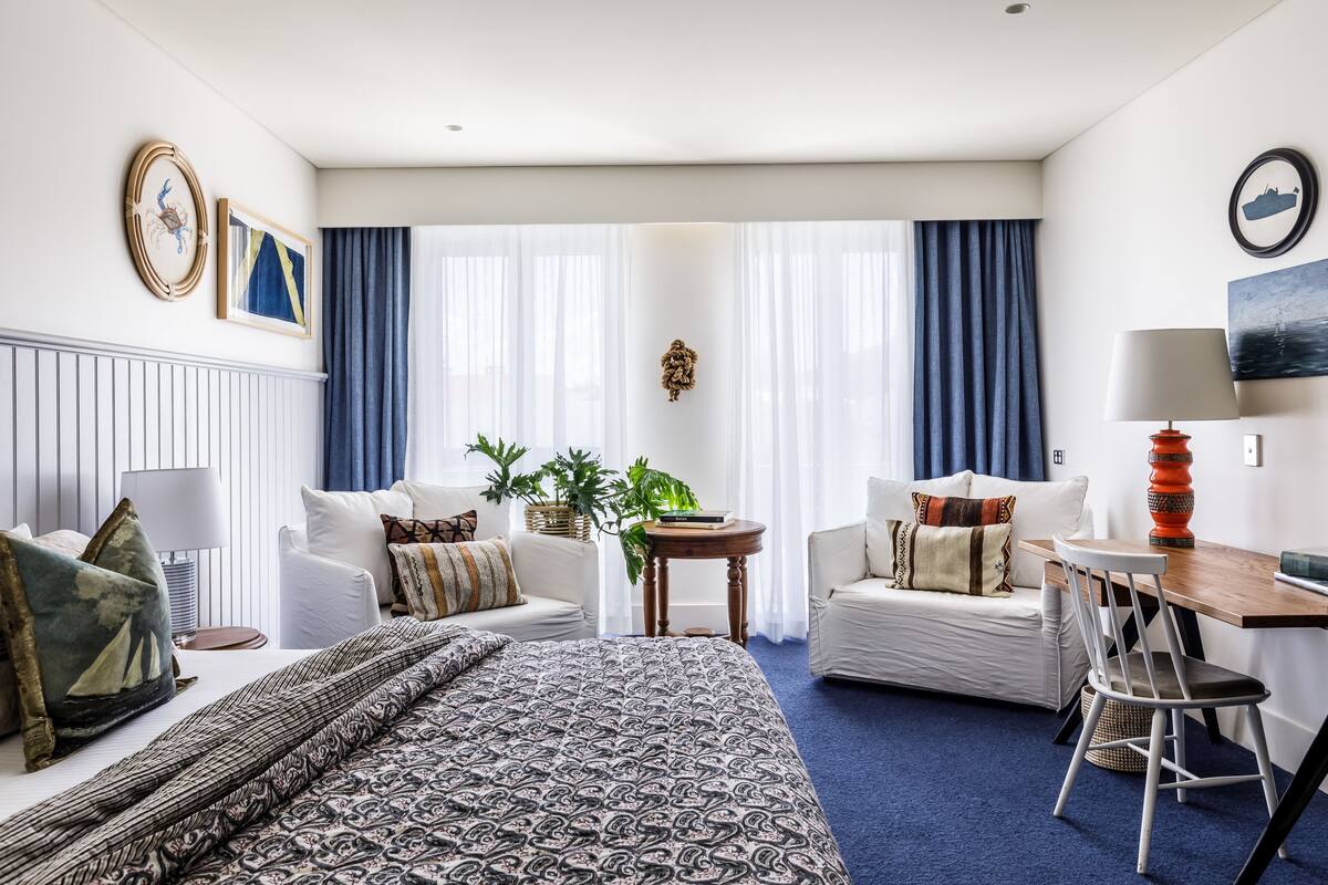 Quarto de hotel moderno com uma cama grande, dois sofás brancos, uma mesa de madeira e uma escrivaninha.