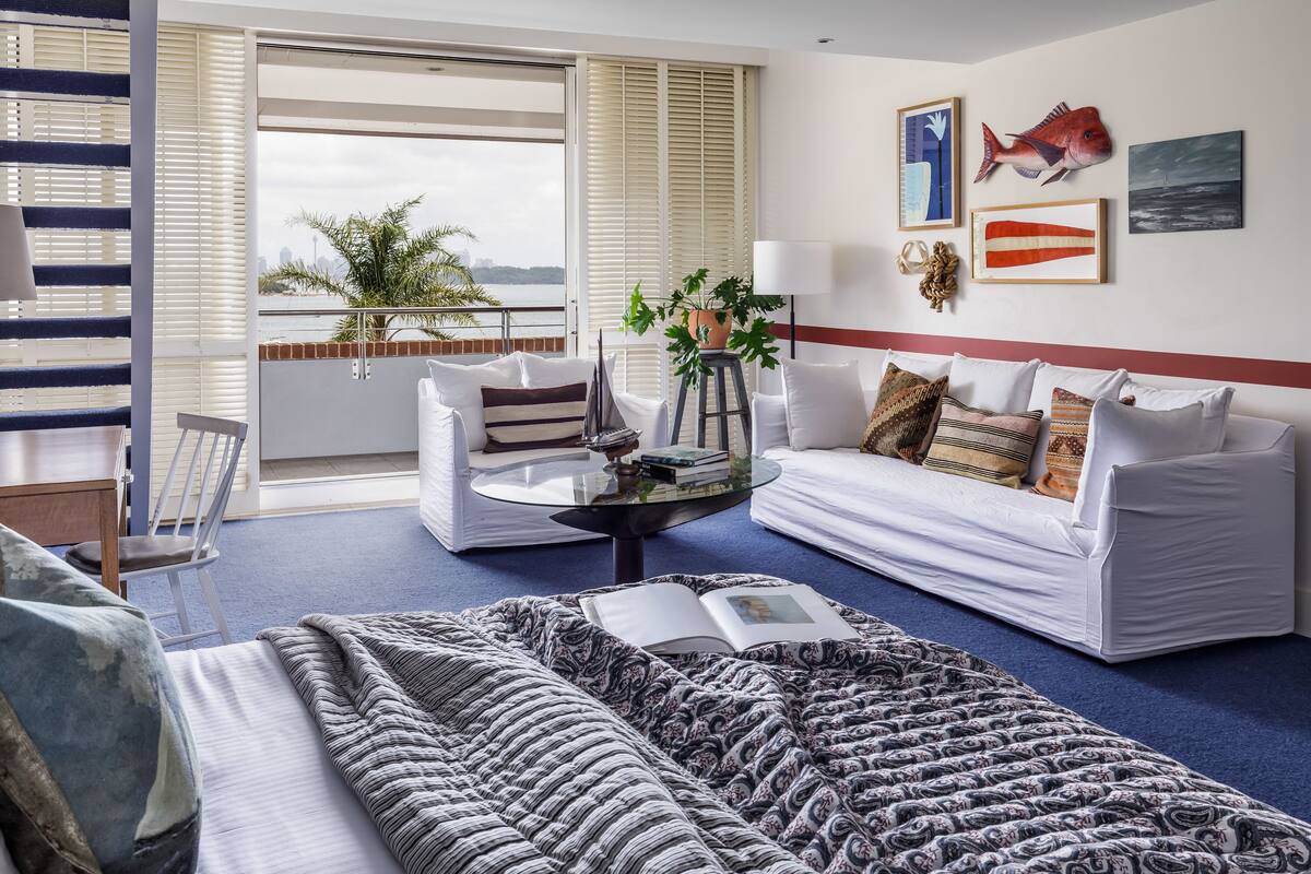 Quarto de hotel moderno com varanda, mobiliário branco, mesa de centro de vidro e vista para o horizonte da cidade.