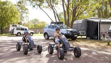 Duas crianças em karts em uma entrada de garagem, com uma van e um carro ao fundo.