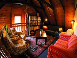 Interior acolhedor de uma cabana de madeira, com teto de madeira, uma estante, um sofá, uma poltrona e uma mesa de centro.