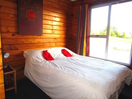 Quarto de cabana de madeira com cama, um banquinho de madeira e uma janela com cortinas.