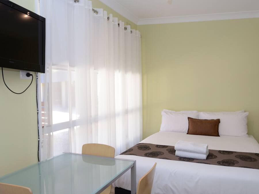 Quarto de hotel com cama, televisão na parede, mesa de vidro e uma cadeira.