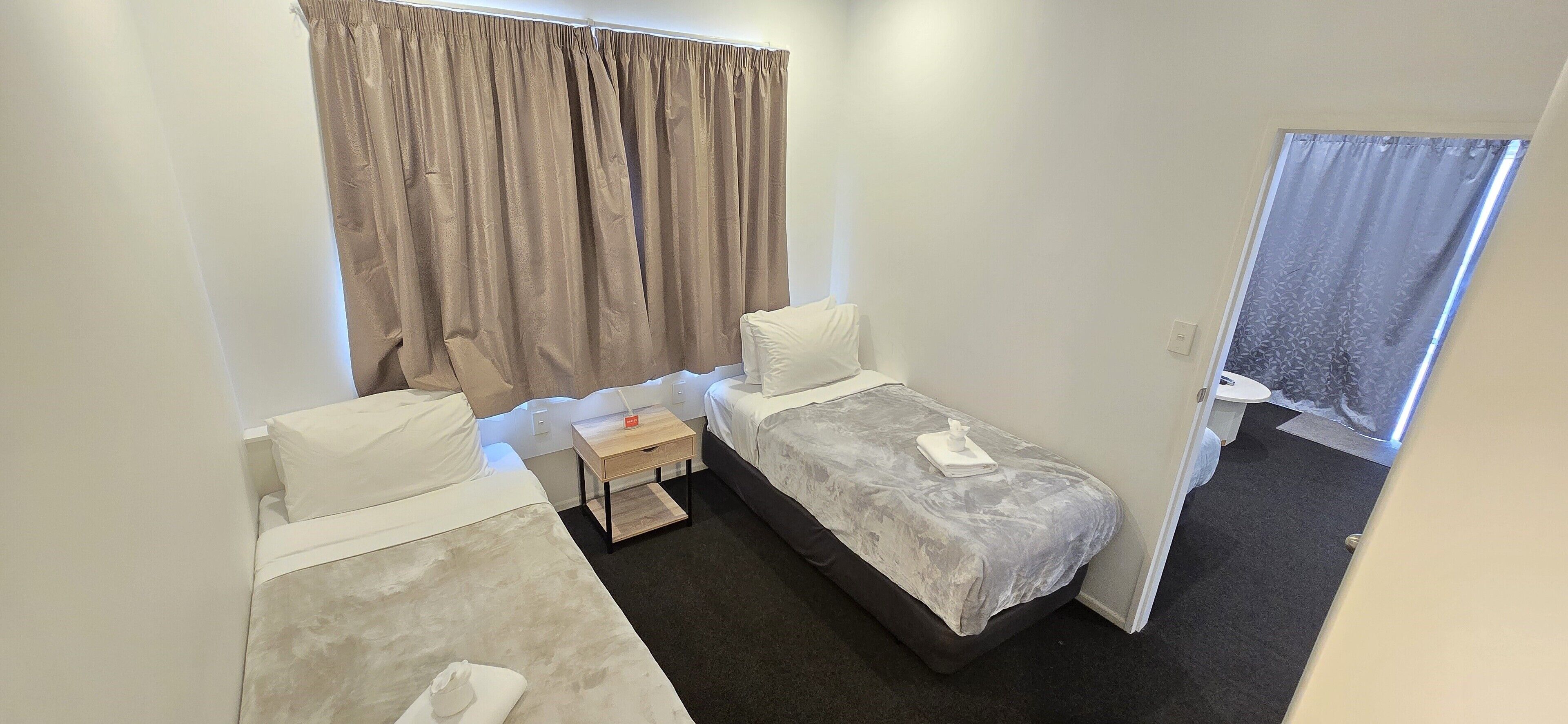 Quarto de hotel com duas camas, uma mesa pequena com abajur, banheiro com vaso sanitário e pia, e uma porta que dá para outro quarto.
