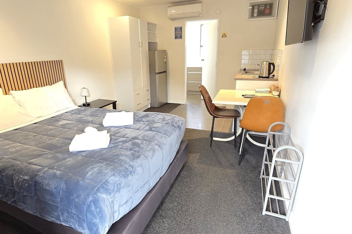 Quarto de hotel com cama, escrivaninha, cadeira, guarda-roupa e cozinha compacta.