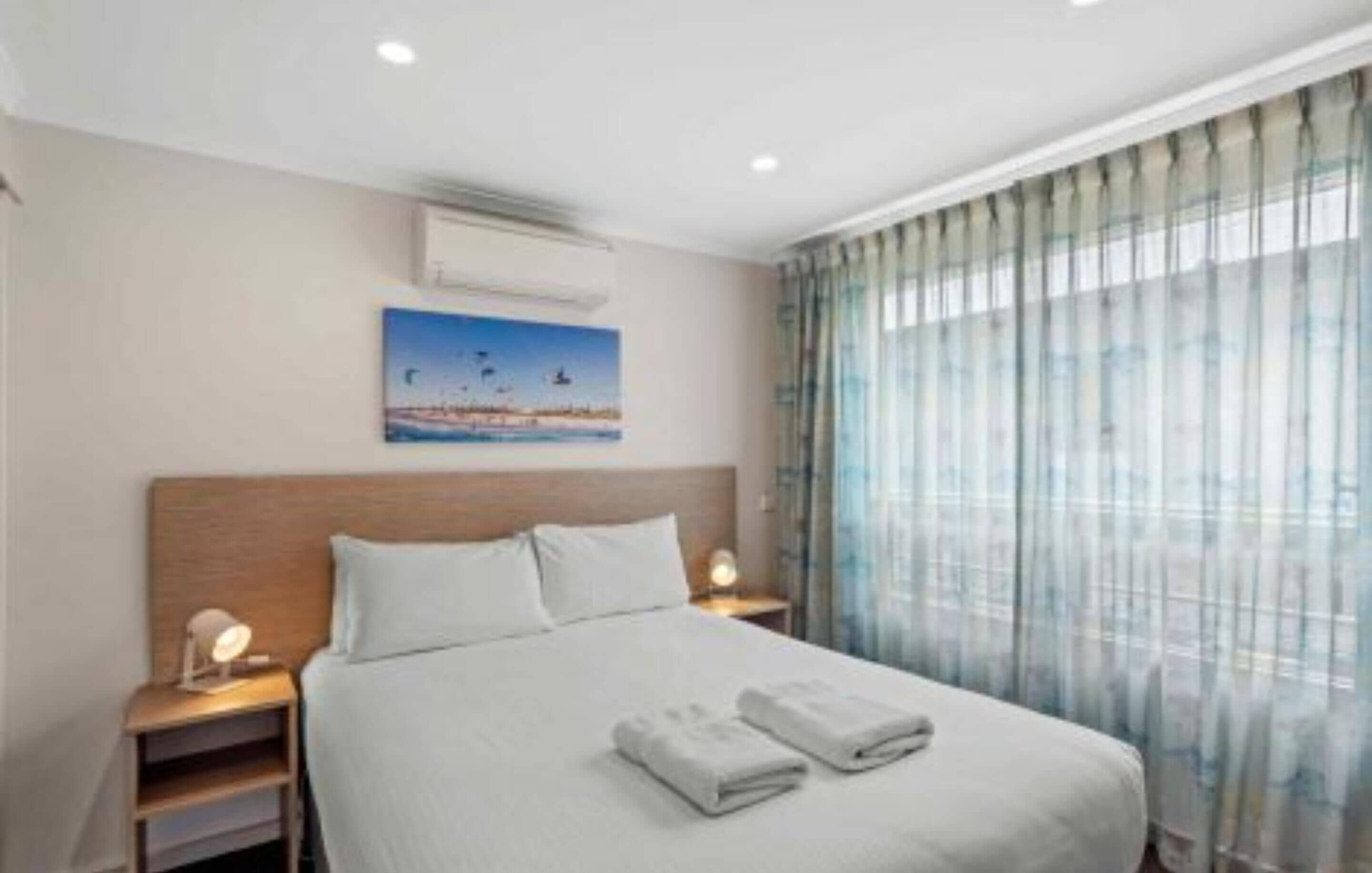 Quarto de hotel com uma cama grande, duas mesas de cabeceira com luminárias, um quadro na parede e vista através de cortinas translúcidas.