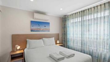 Quarto de hotel com uma cama grande, duas mesas de cabeceira com luminárias, um quadro na parede e vista através de cortinas translúcidas.