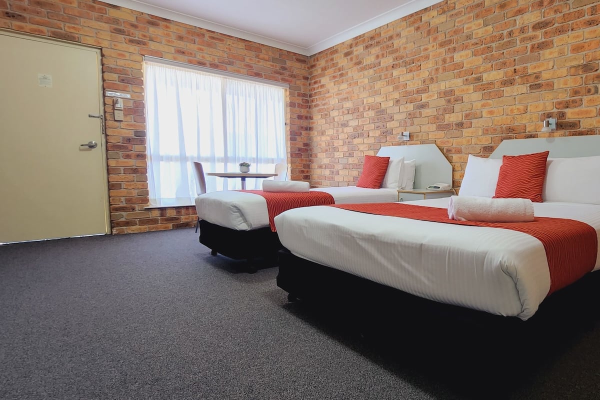 Quarto de hotel com duas camas, uma parede de tijolos, uma janela com cortinas, uma mesa pequena e uma cadeira.