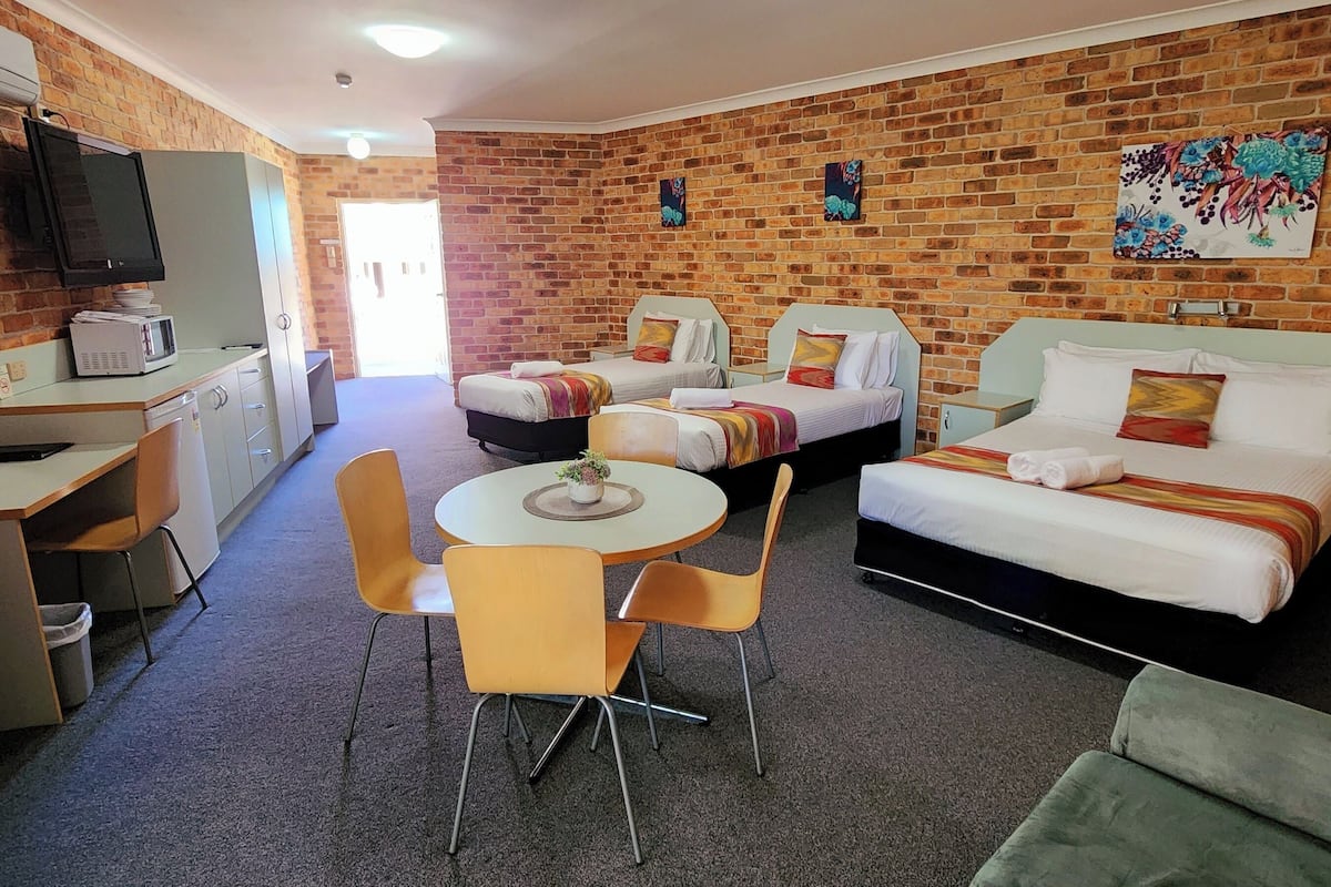Quarto de hotel com parede de tijolos, duas camas, área de refeições com cadeiras, pequena cozinha e televisão.