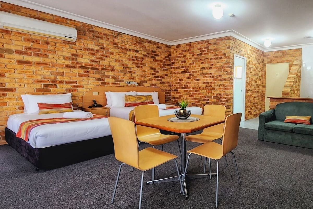 Quarto de hotel com paredes de tijolo, duas camas, uma mesa de jantar com cadeiras e um sofá verde.