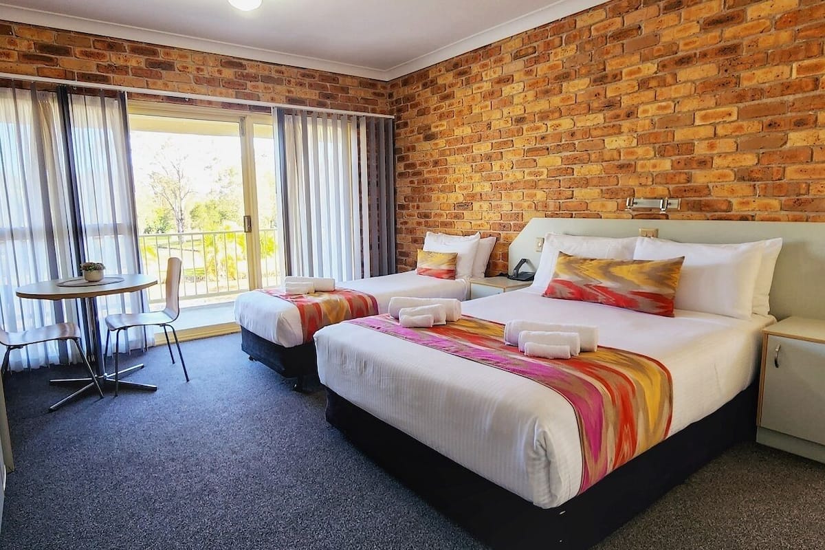 Quarto de hotel com parede de tijolos, duas camas, uma mesa pequena e uma cadeira.