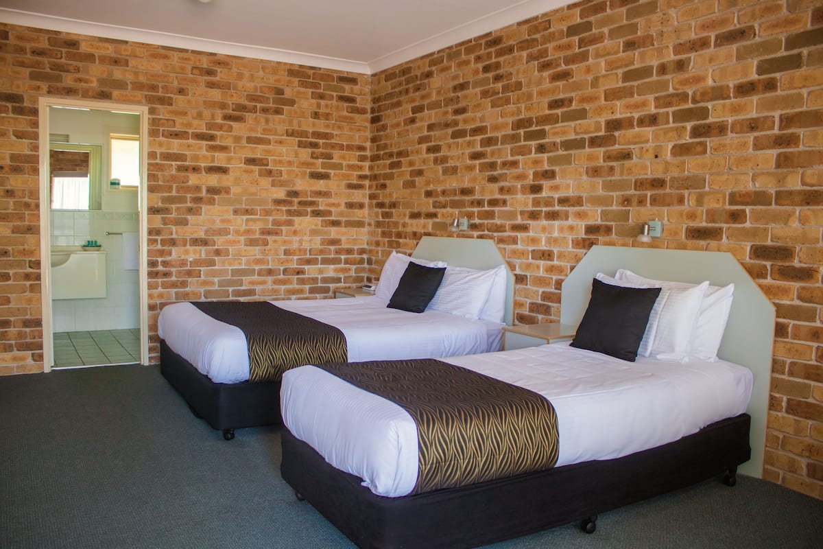 Quarto de hotel com duas camas, uma parede de tijolos e um banheiro visível através de uma porta aberta.