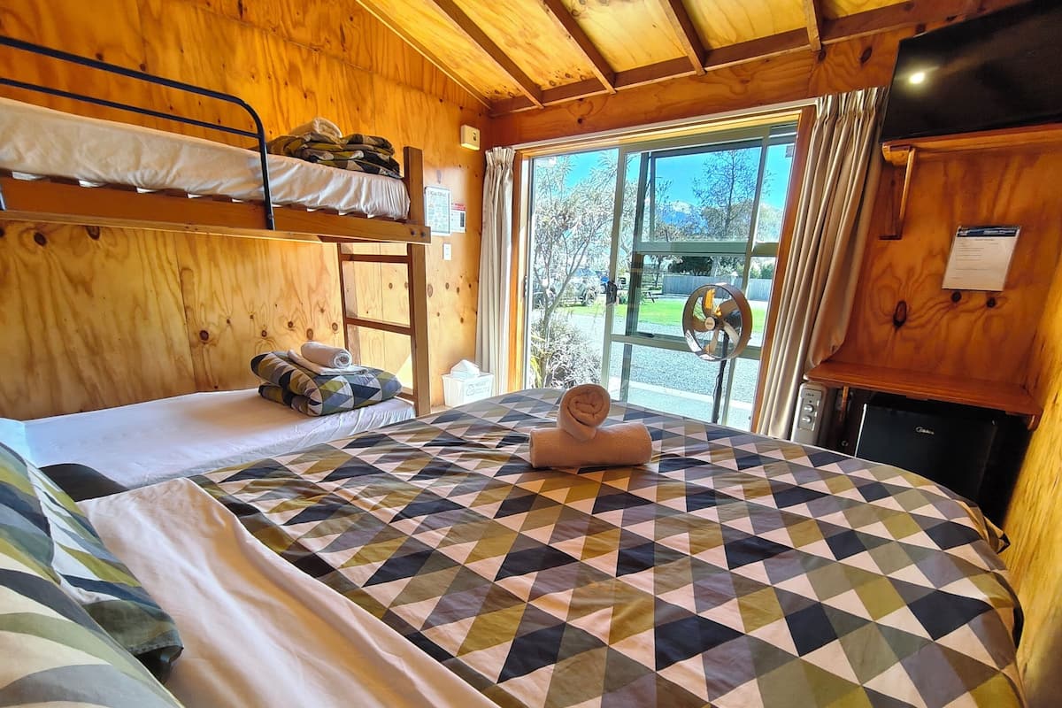 Quarto com duas camas, incluindo uma beliche, paredes de madeira, janela com cortinas e um ventilador.