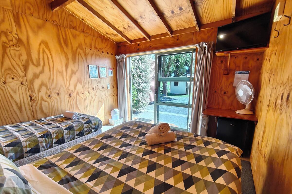 Quarto de cabana de madeira com duas camas, uma janela e um ventilador.