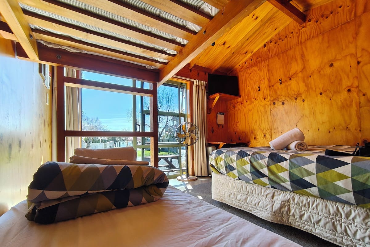 Um quarto de cabana aconchegante com uma cama, um banquinho com almofadas, um ventilador e vista para o exterior.