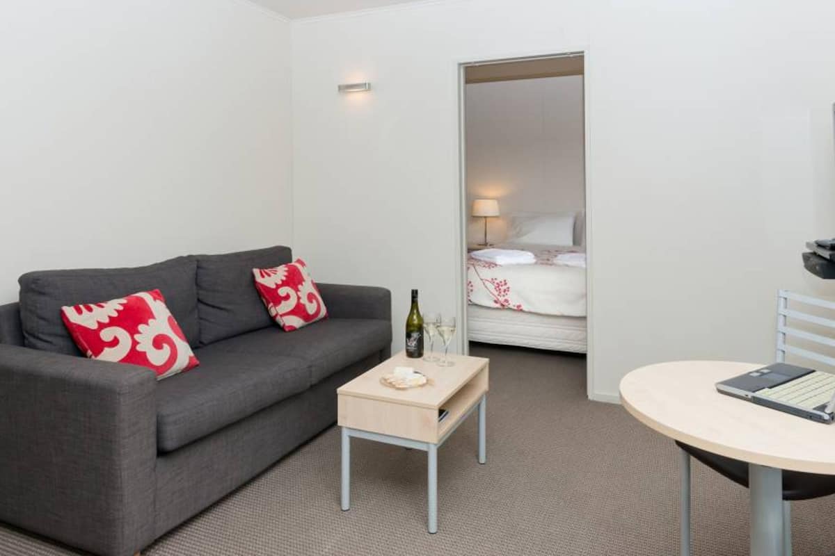 Quarto de hotel compacto com sofá cinza, uma pequena mesa lateral de madeira, uma mesa redonda com um laptop e uma cama com colcha floral rosa e branca.