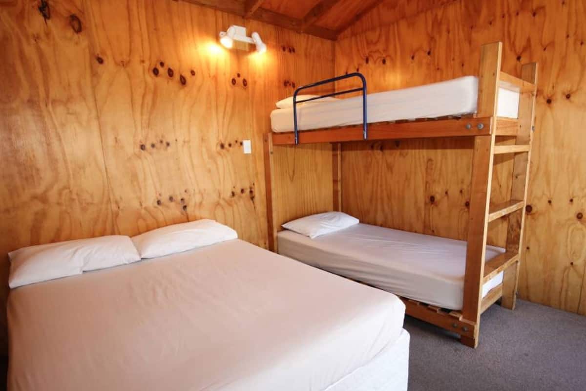 Quarto de cabana de madeira com beliches, uma escada e luminária fixada na parede.