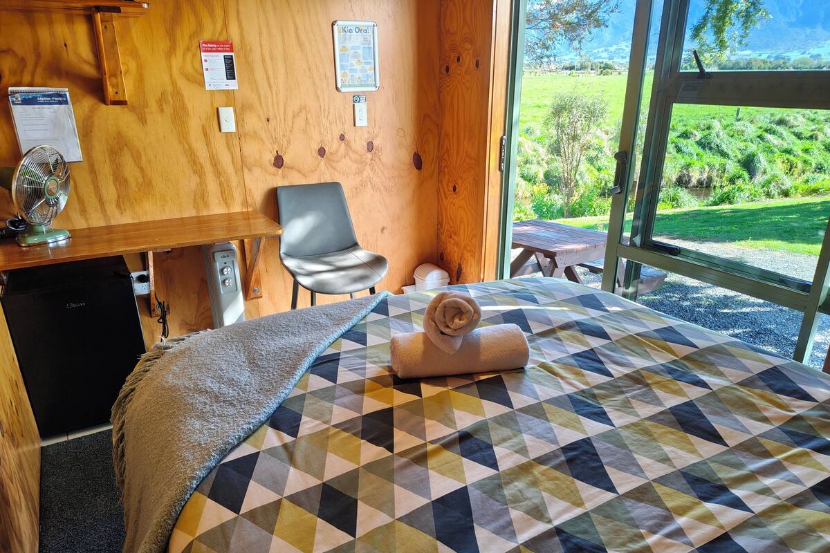 Quarto com cama, cadeira, escrivaninha, ventilador e vista para o exterior.