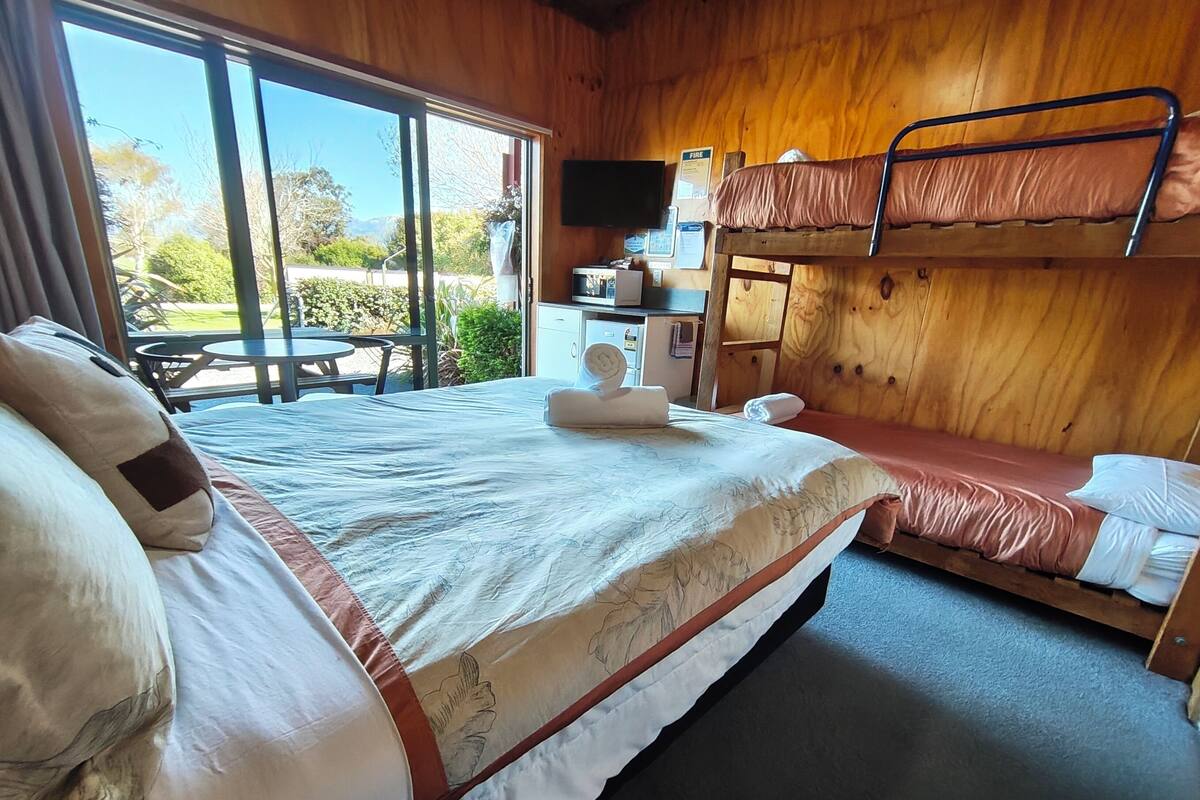 Quarto de hotel com uma cama grande, uma cama beliche menor, uma mesa com cadeira e vista para o exterior por uma porta de correr.