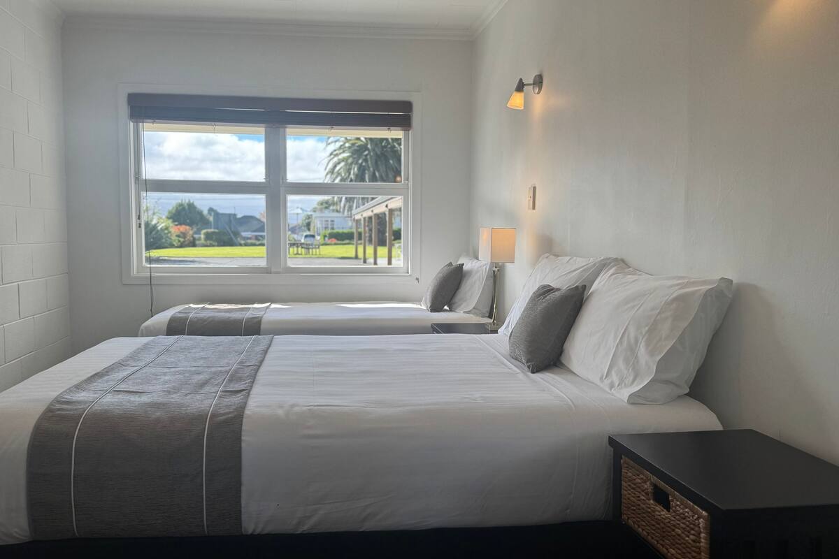 Quarto com duas camas, uma janela ampla com vista para o exterior, mesa de cabeceira com luminária e um criado-mudo.