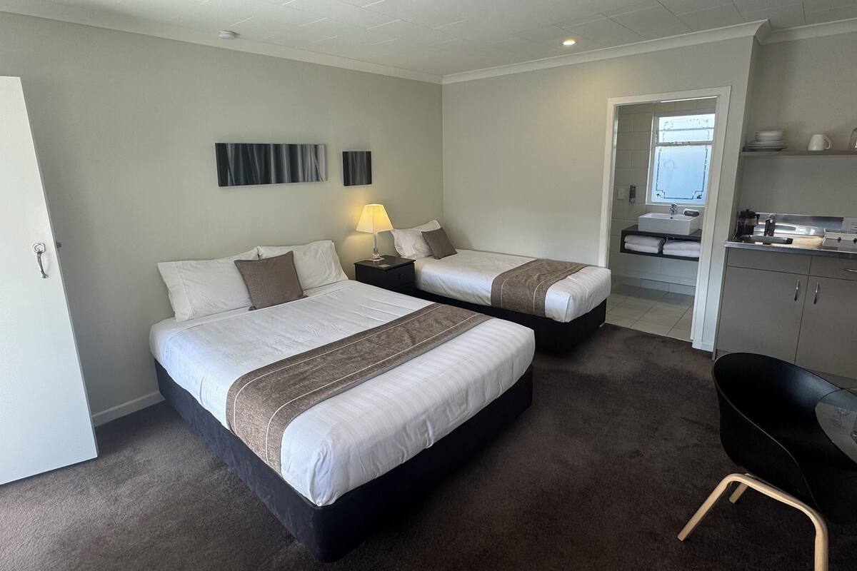 Quarto de hotel com duas camas, uma escrivaninha, uma cadeira, uma pequena cozinha e um banheiro visível por uma porta aberta.