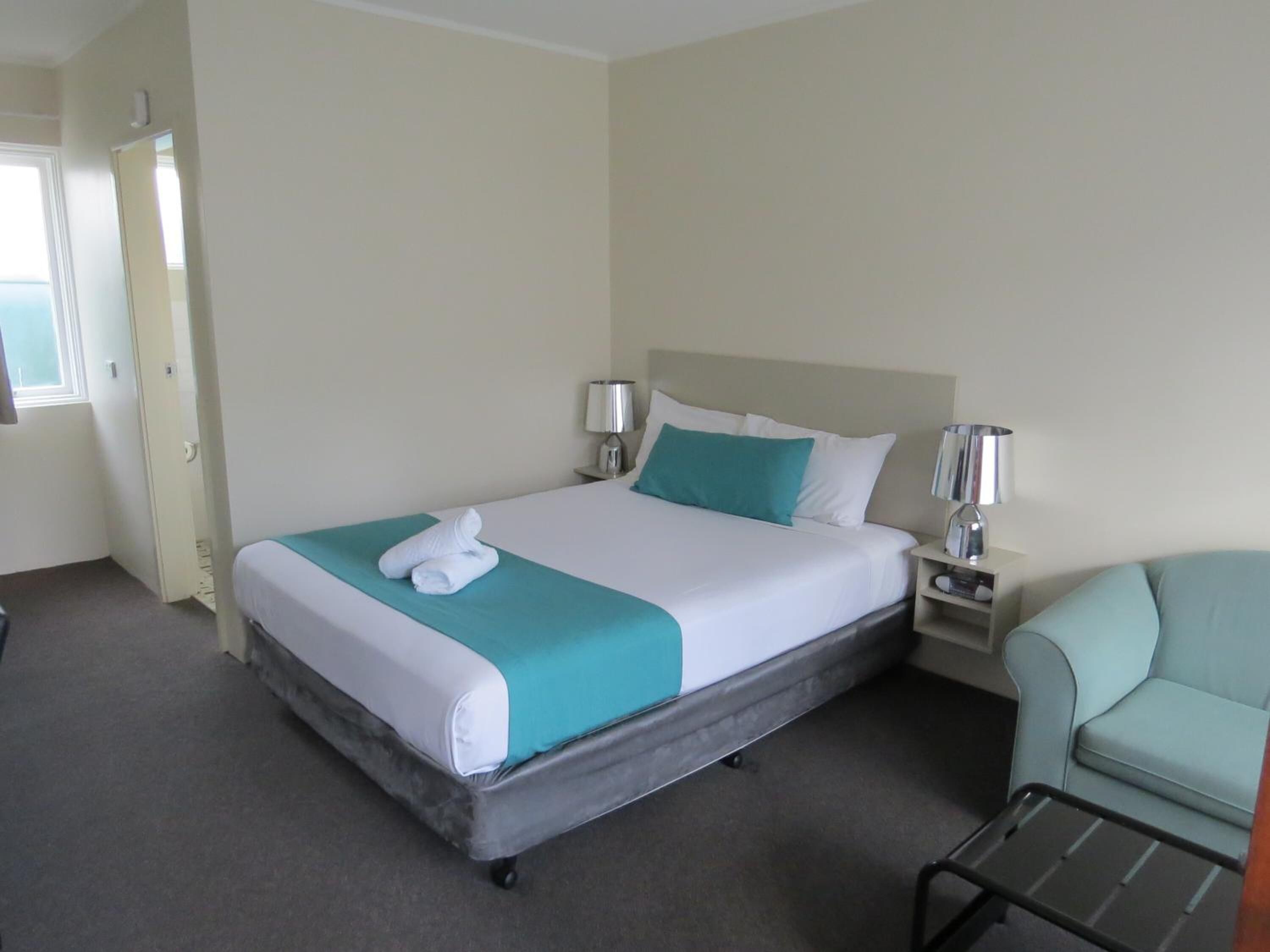 Quarto de hotel com uma cama, duas luminárias de cabeceira, uma cadeira e uma pequena mesa.