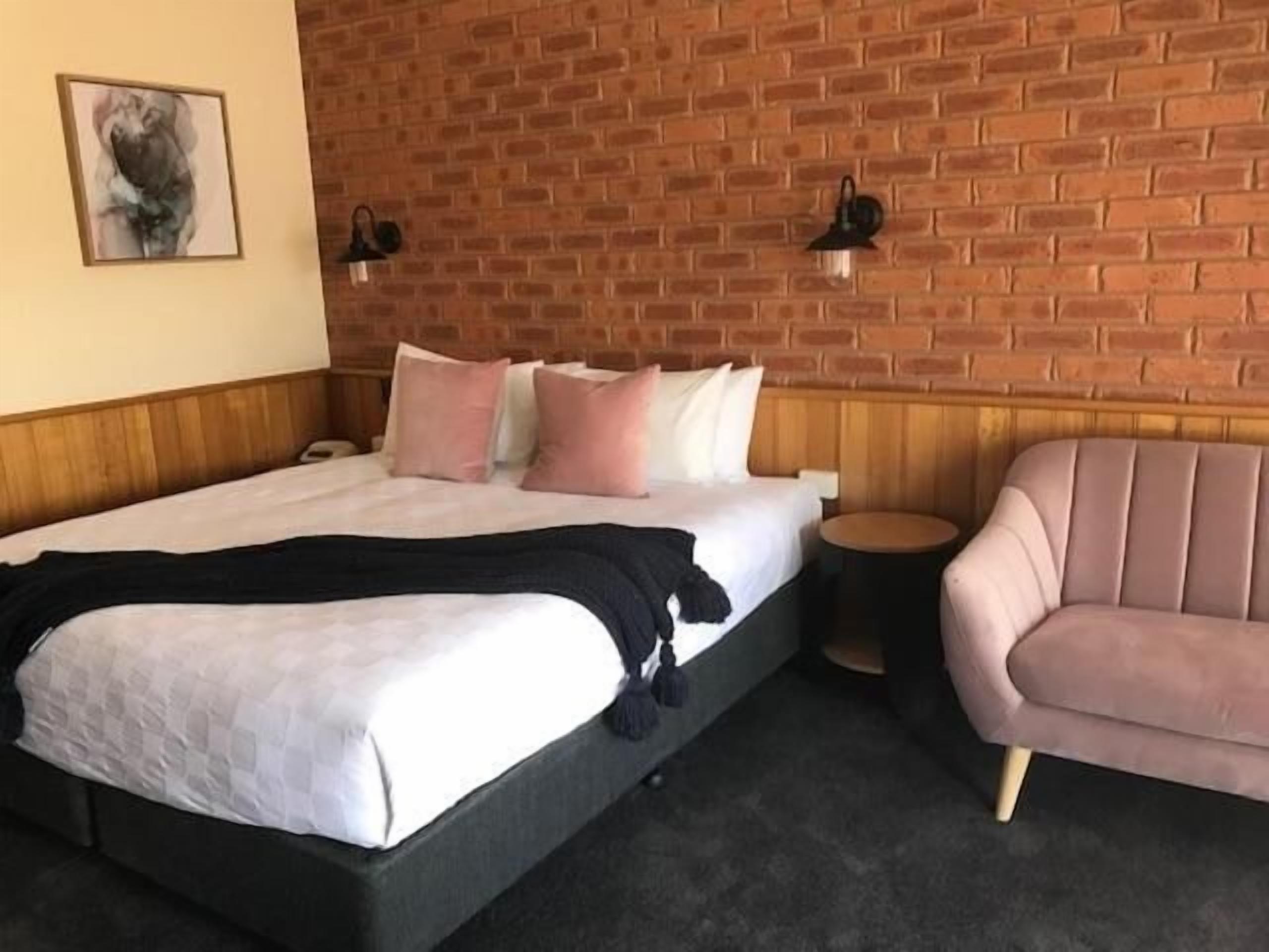 Quarto de hotel com parede de tijolos, uma cama com roupa de cama branca, um cobertor preto e uma cadeira rosa, além de duas luminárias fixadas na parede.
