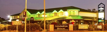 Um hotel com fachada iluminada em verde, um letreiro para o drive-thru 'Cellarbractions' e um anúncio com a palavra 'ALMOÇO' e o preço de $10.90.