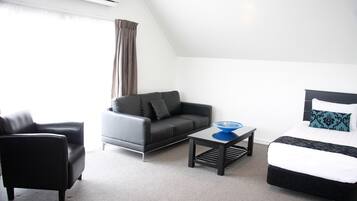 Quarto de hotel moderno com um sofá de couro preto, uma pequena mesa com uma tigela azul, uma cama com cabeceira preta e uma janela com cortinas.