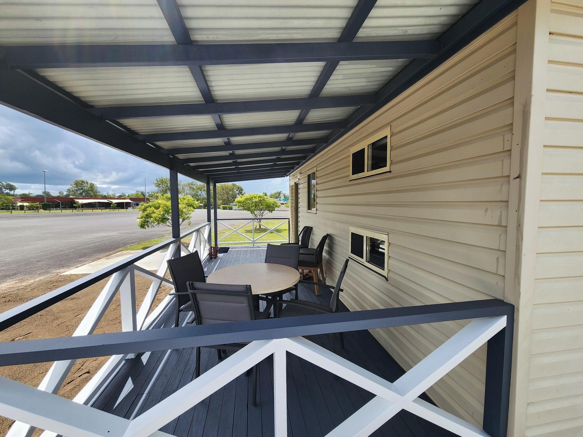 Uma área de estar coberta ao ar livre, com mesa, cadeiras, guarda-corpo de metal e vista para uma estrada e árvores.