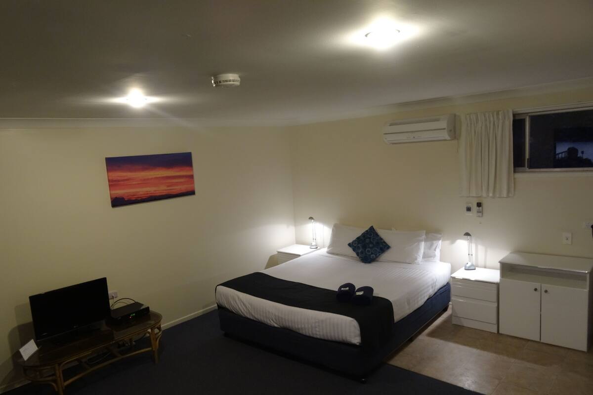Quarto de hotel com uma cama, luminárias de cabeceira, escrivaninha, televisão e um quadro na parede.