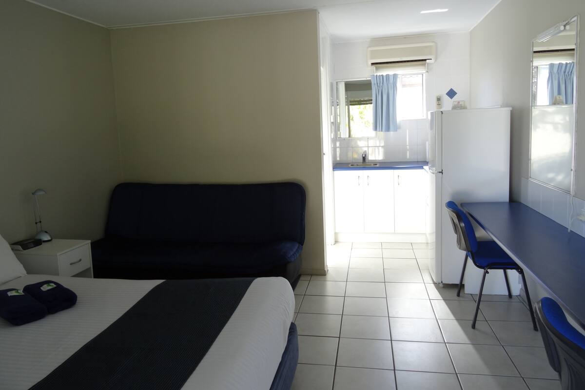 Quarto de hotel com cama, escrivaninha, cadeira, pequeno kitchenette e banheiro.