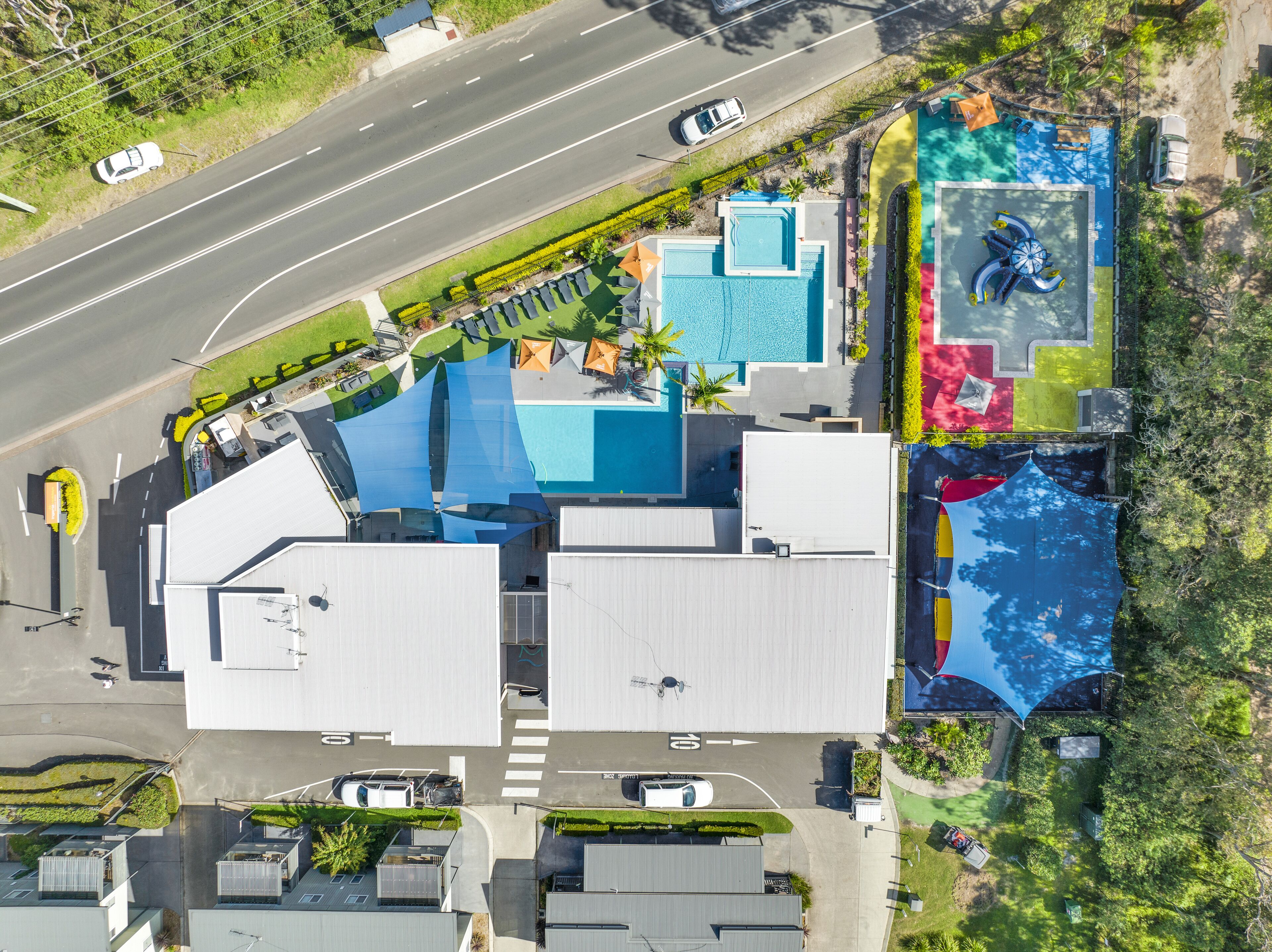 Vista aérea de um edifício moderno com piscina, várias vagas de estacionamento e uma estrada adjacente à propriedade.