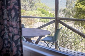 Uma mesa de madeira e uma cadeira em frente a um prédio, com vista para vegetação e colinas ao fundo.