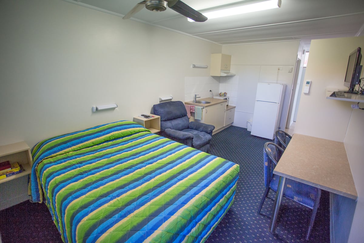 Quarto com cama, escrivaninha, cadeira, cozinha compacta e frigorífico.