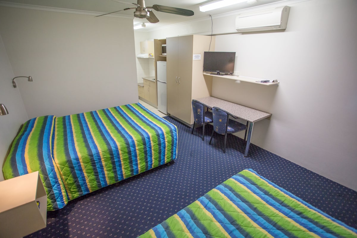 Quarto de hotel com duas camas, uma escrivaninha, uma cadeira, uma televisão e um ventilador de teto.