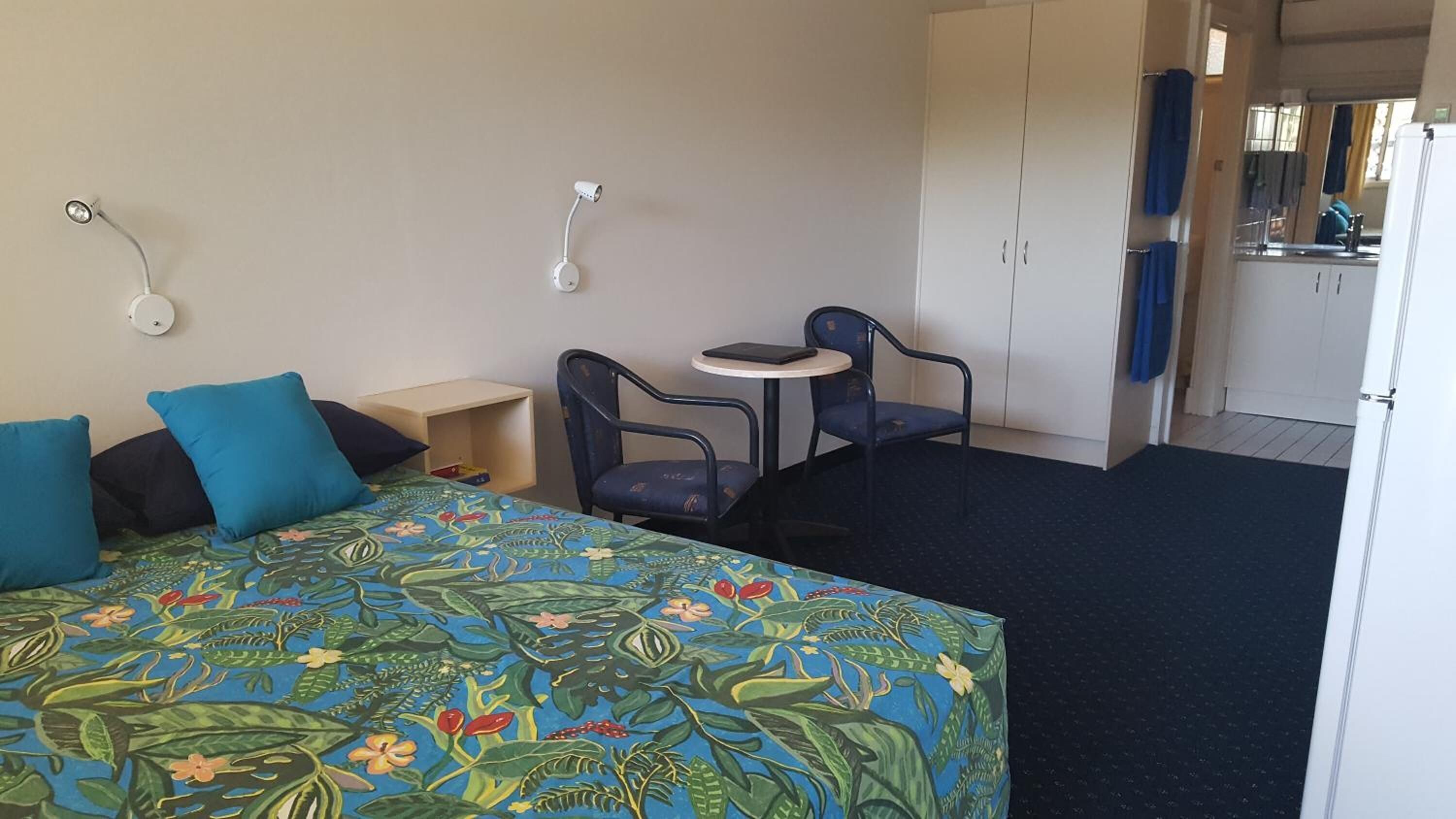 Quarto de hotel com cama, duas cadeiras, uma mesinha, abajur, guarda-roupa e banheiro visível através de uma porta aberta.