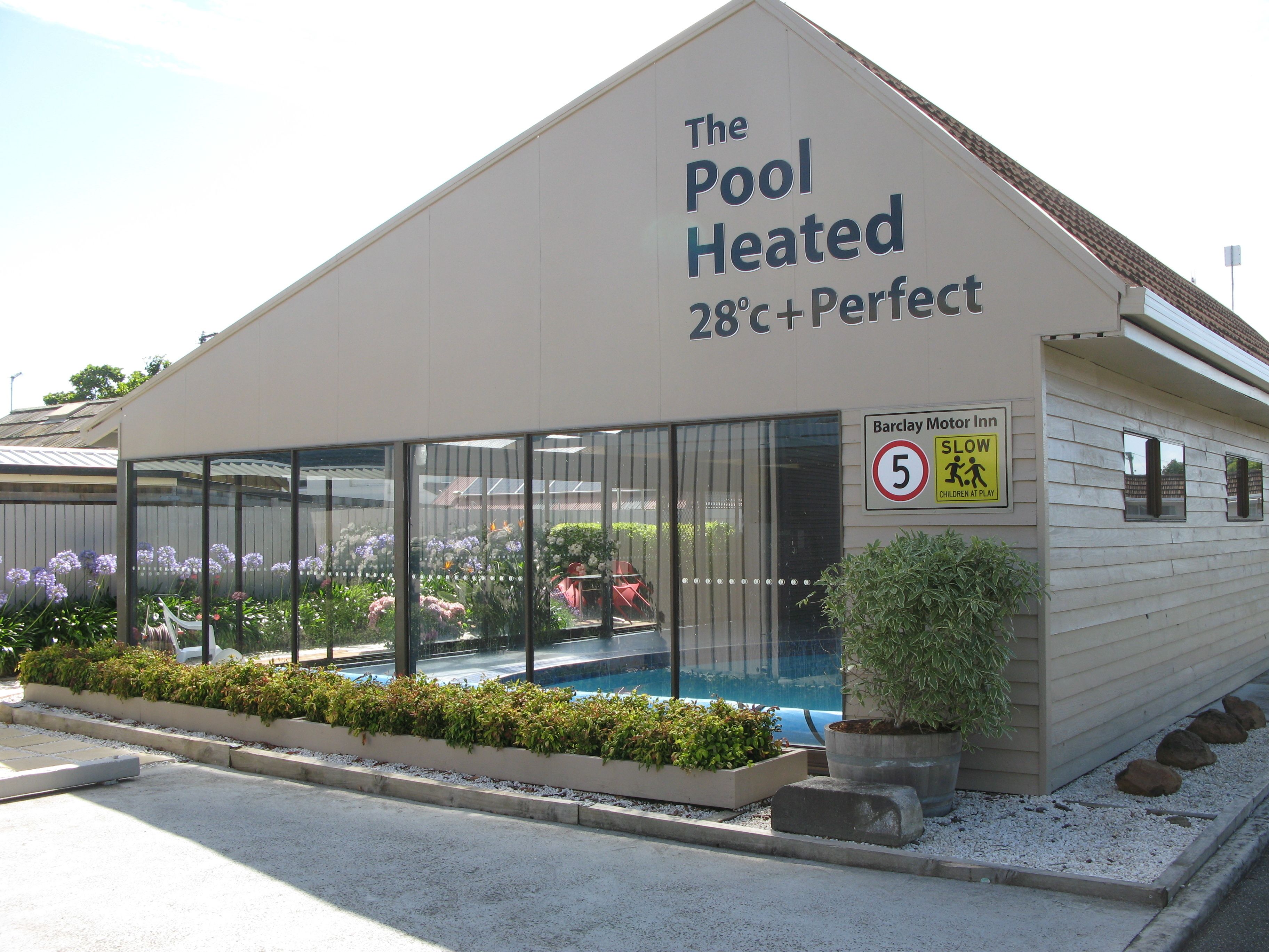 Um edifício com a placa "The Pool Heated 28°C + Perfect" e uma placa indicando "Barclay Motor Inn", além de uma placa de limite de velocidade de "5".