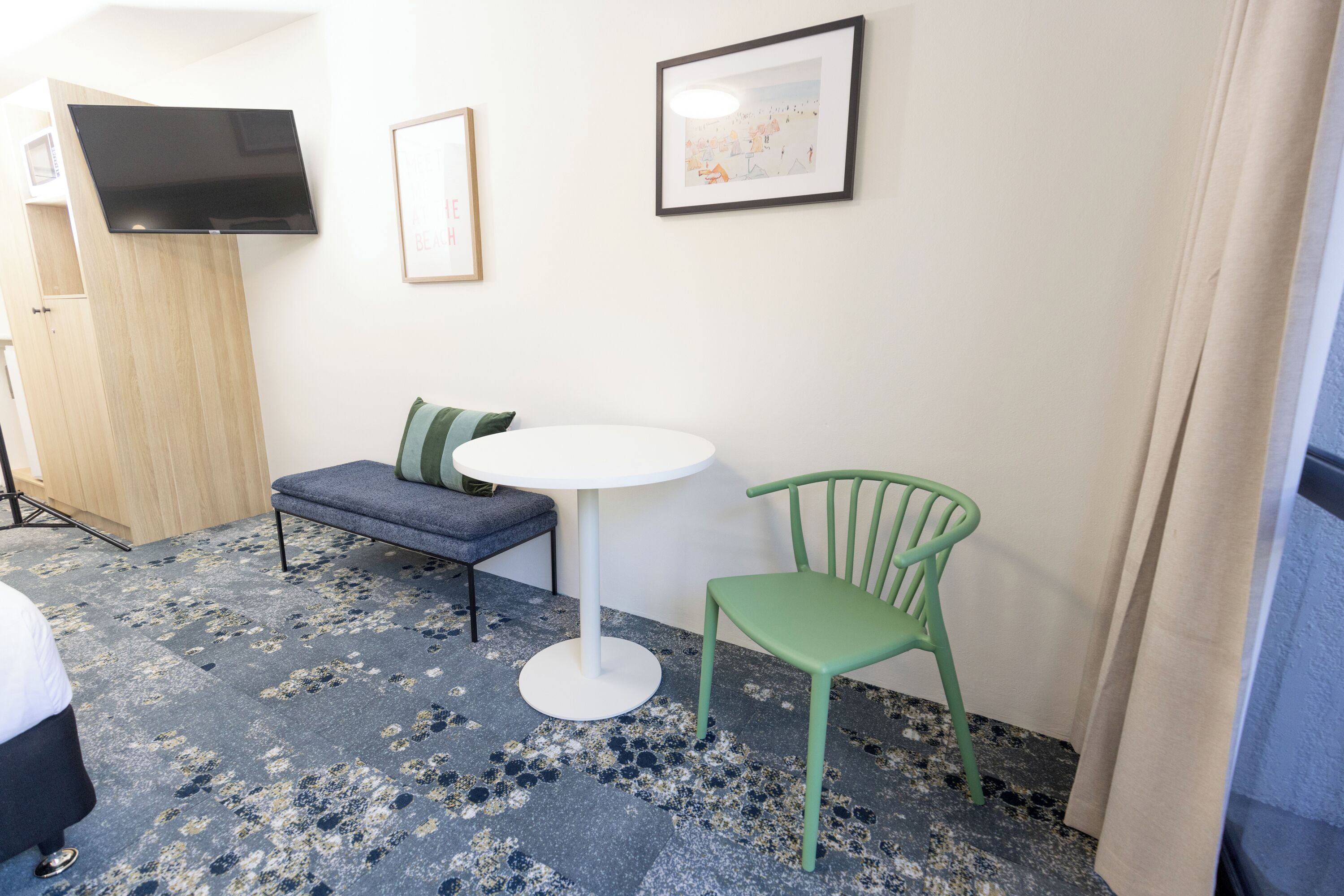 Um quarto pequeno com uma mesa redonda branca, uma cadeira verde, um sofá cinza com uma almofada verde, uma televisão de tela plana na parede e um quadro emoldurado.
